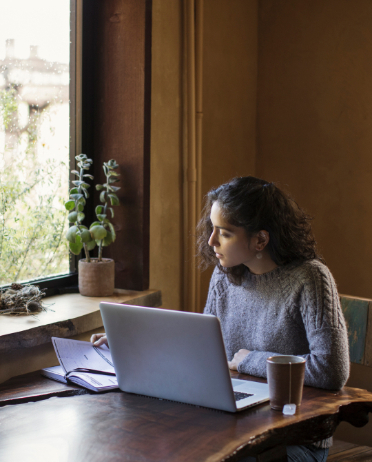 A woman glances out a window while sitting at a table with a laptop and ...
